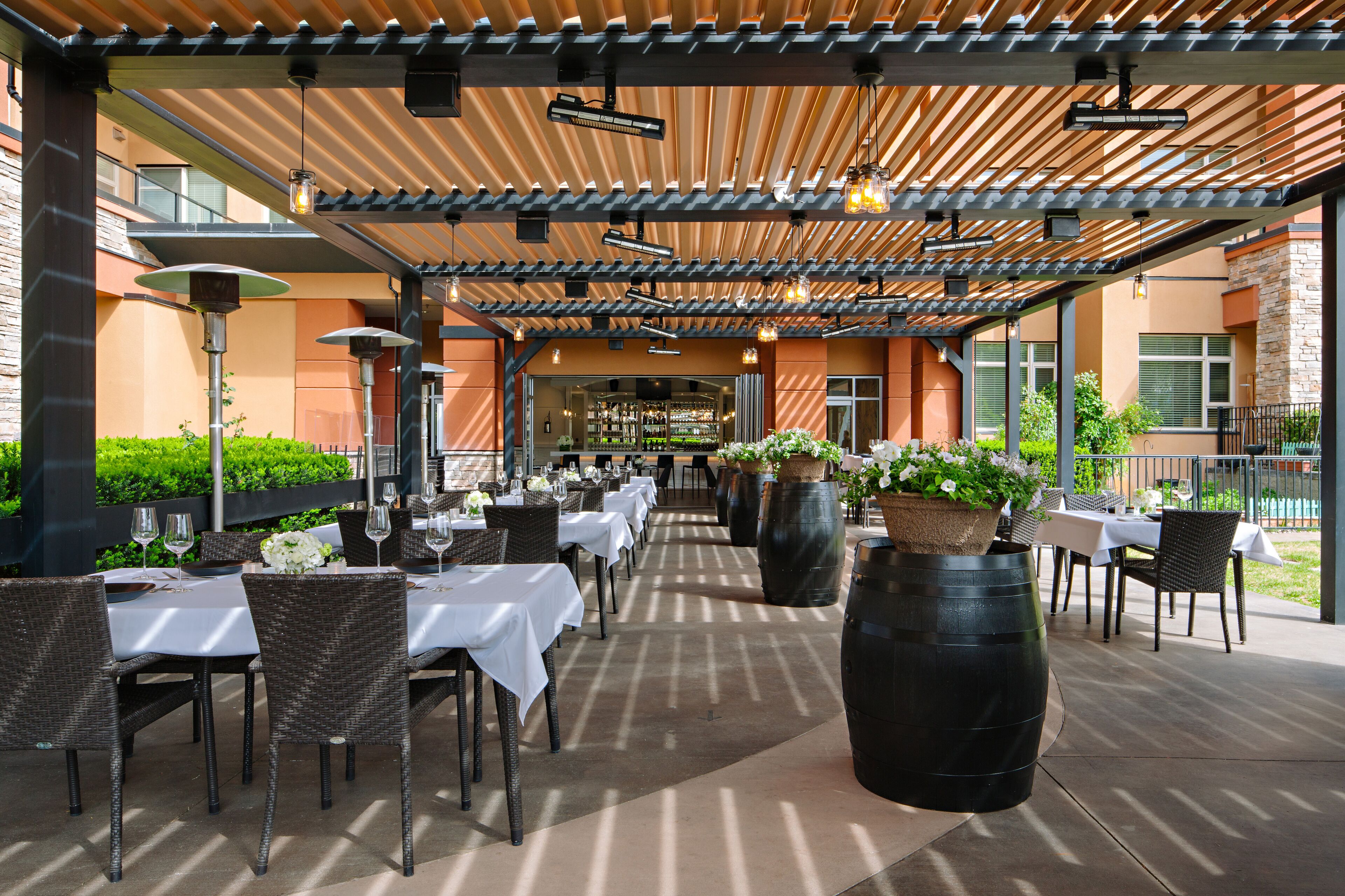 Uma área de refeições ao ar livre com mesas e cadeiras, uma pérgola com luzes penduradas e plantas em vasos.
