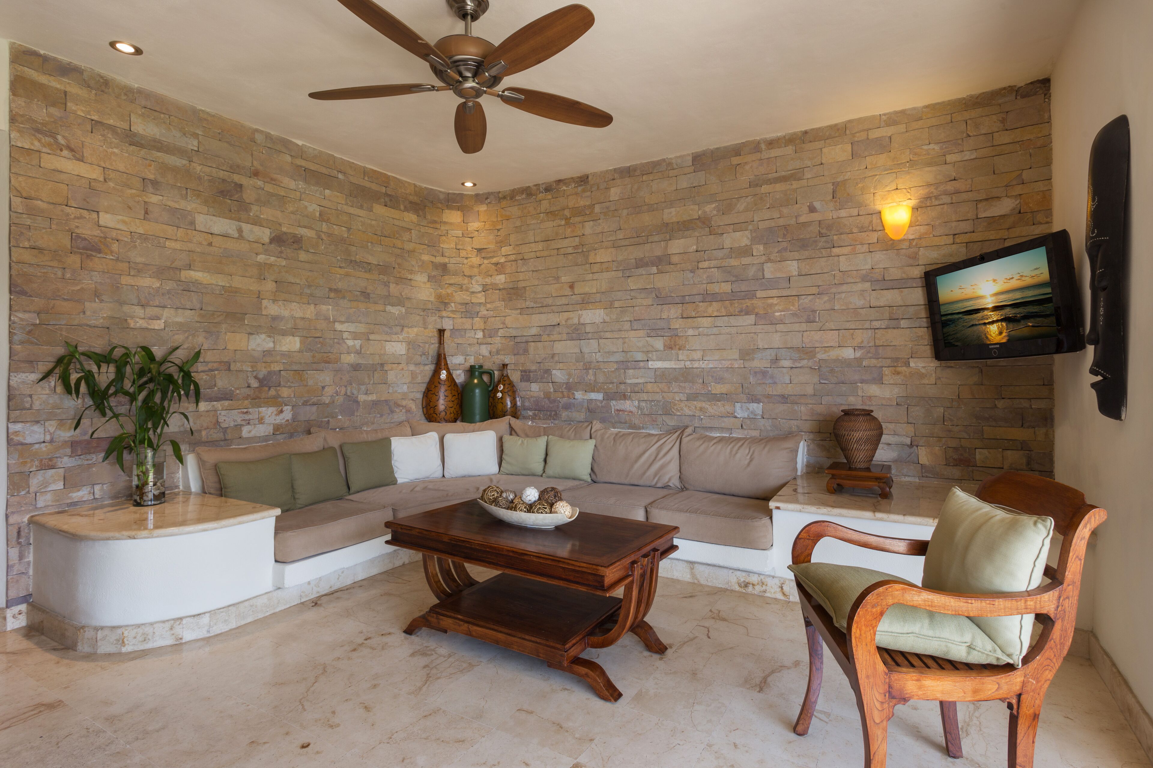 Uma sala de estar com parede de pedra, um ventilador de teto, uma TV montada na parede, um sofá, uma mesa de centro e uma cadeira de madeira.