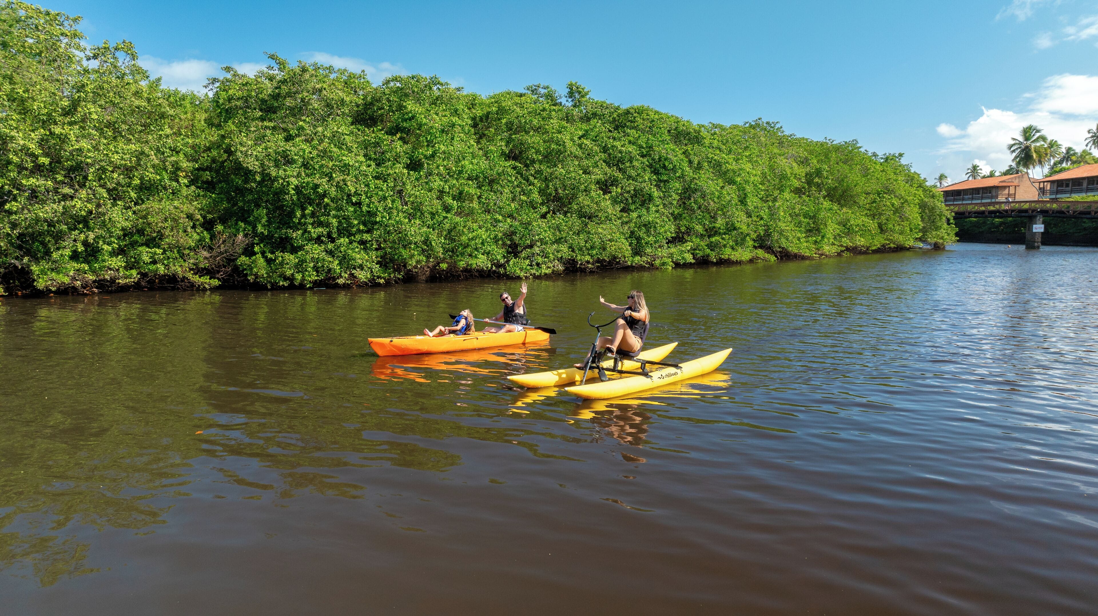 Três pessoas em canoas em um rio calmo, cercadas por vegetação exuberante e céu claro.