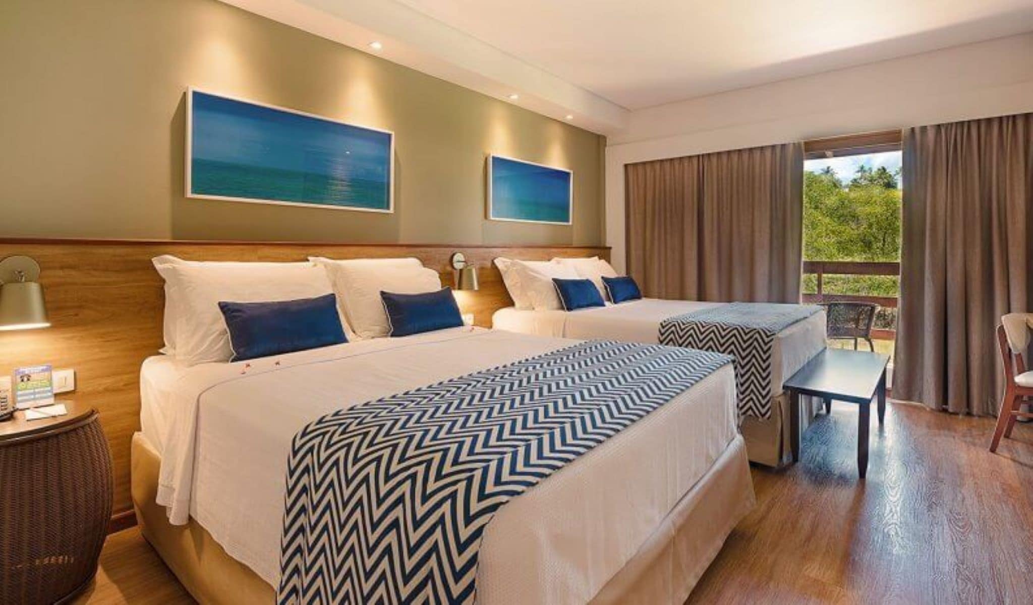 Quarto de hotel com duas camas, uma mesinha de cabeceira, uma mesa e vista para o exterior.