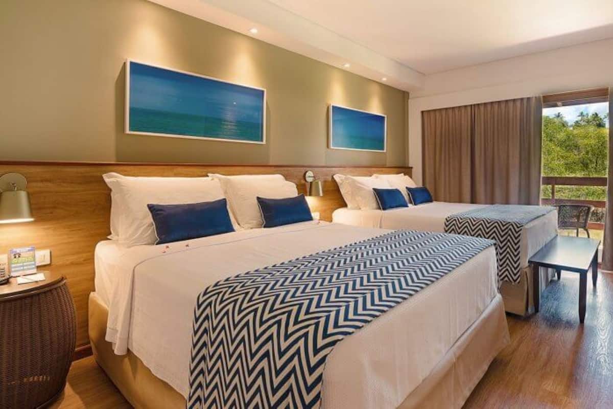 Quarto de hotel com duas camas, uma mesinha de cabeceira, uma mesa e vista para o exterior.