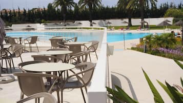 Uma área à beira da piscina com mesas e cadeiras, piscina e palmeiras.
