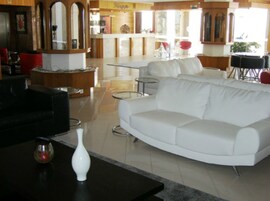 Um lobby de hotel moderno com um sofá branco, uma mesa redonda pequena e uma área de bar ao fundo.