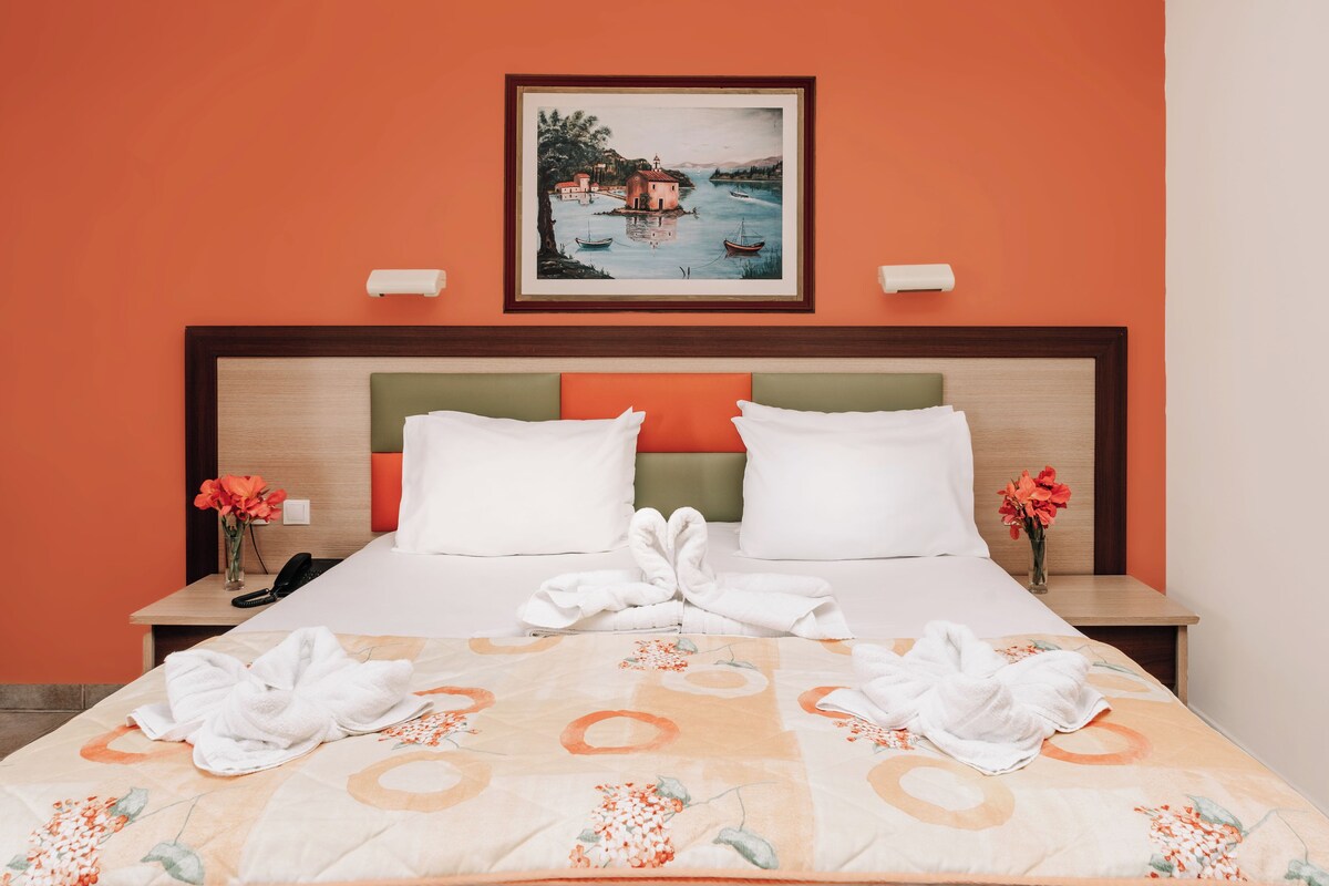 Quarto de hotel com uma cama, duas almofadas, um quadro acima da cama e um vaso com flores na mesa de cabeceira.
