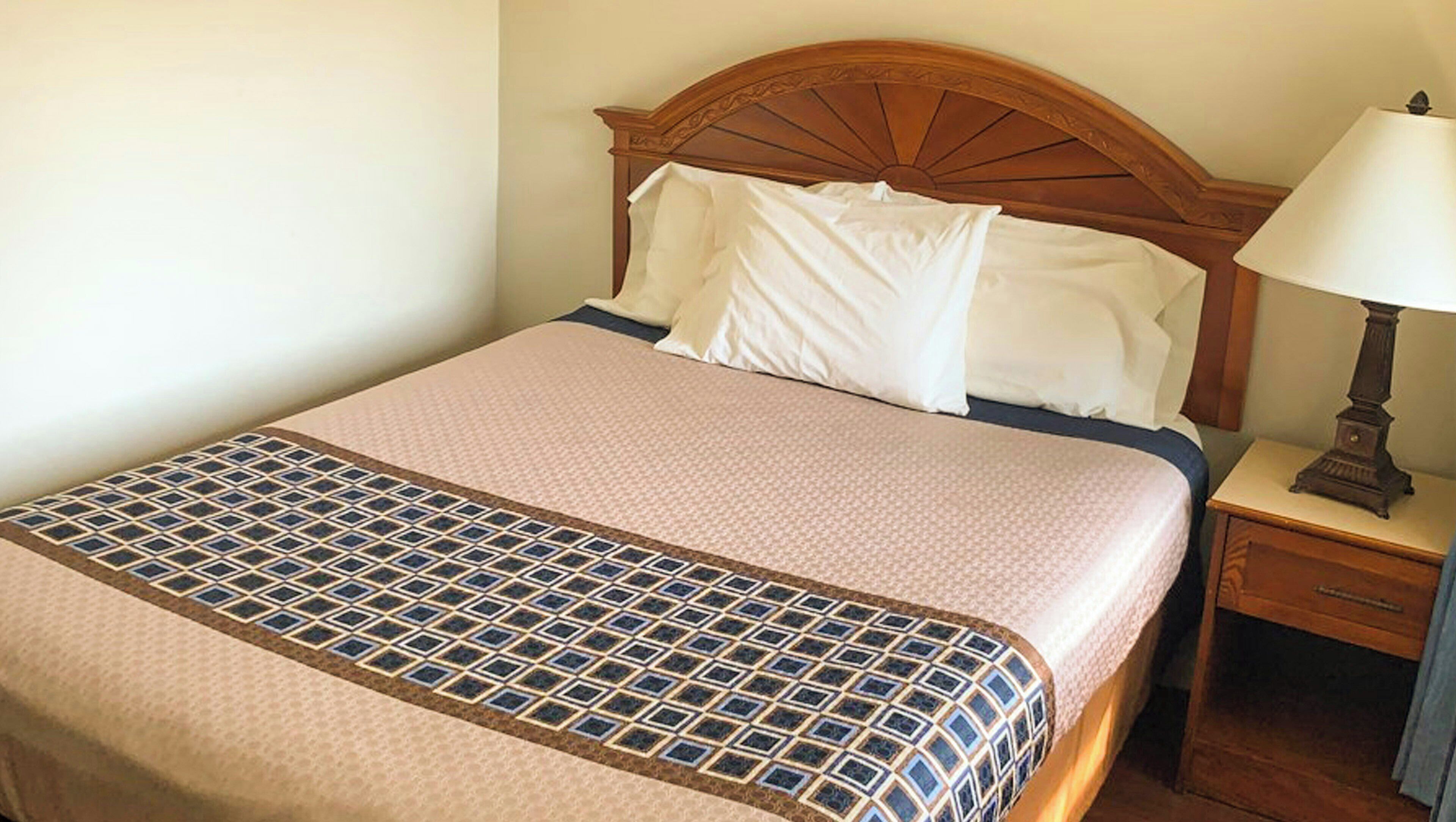 Uma cama de solteiro com colcha estampada, duas almofadas, cabeceira de madeira, mesa de cabeceira com abajur e criado-mudo.