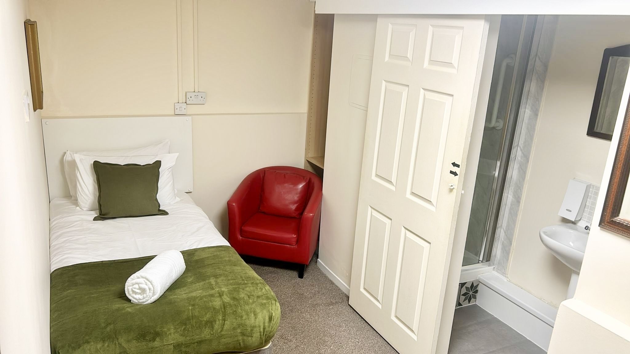 Quarto de hotel com cama, uma cadeira vermelha, banheiro com pia e espelho, e porta que dá para outro cômodo.