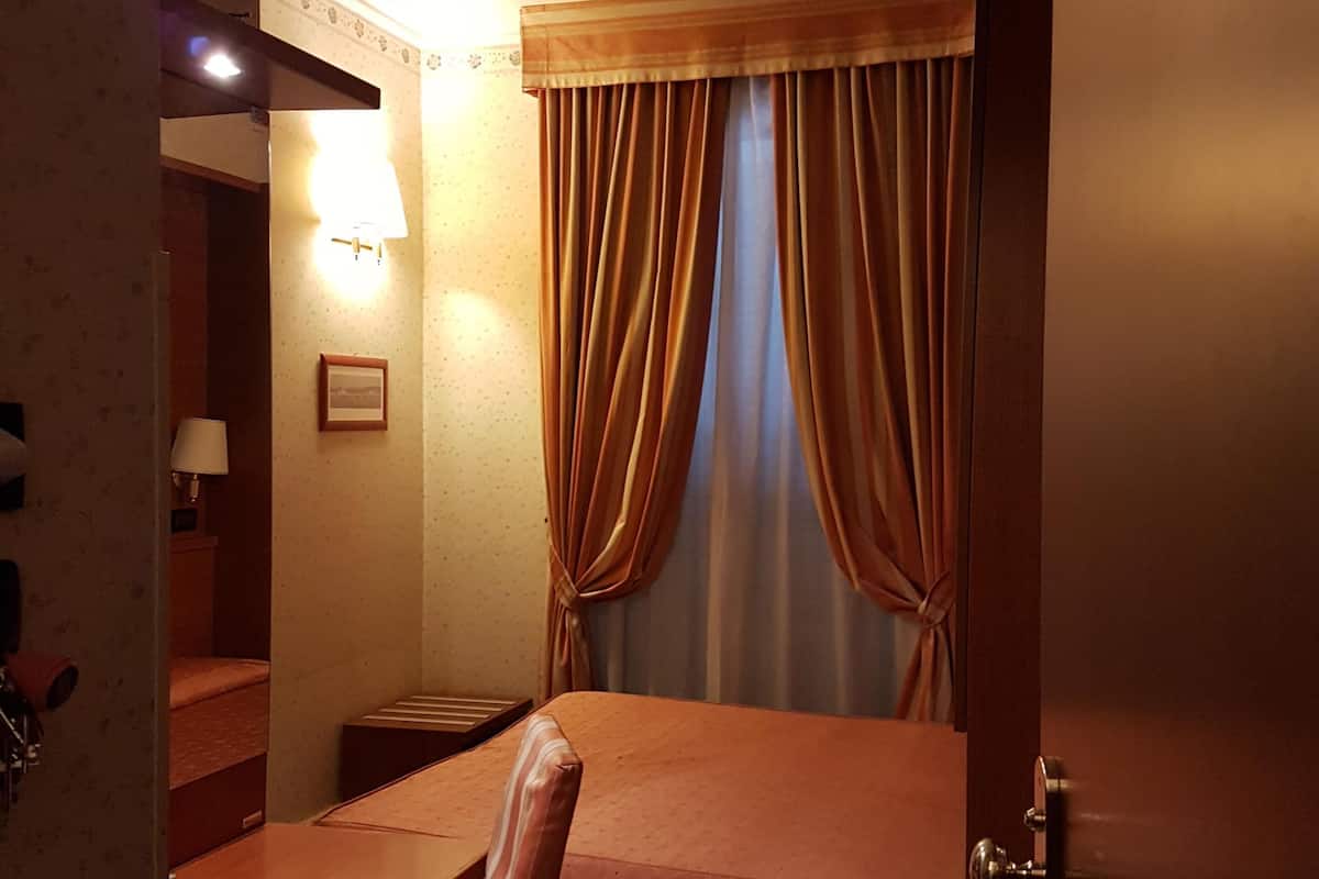 Quarto de hotel com cama, escrivaninha, cadeira, luminária e janela com cortinas.