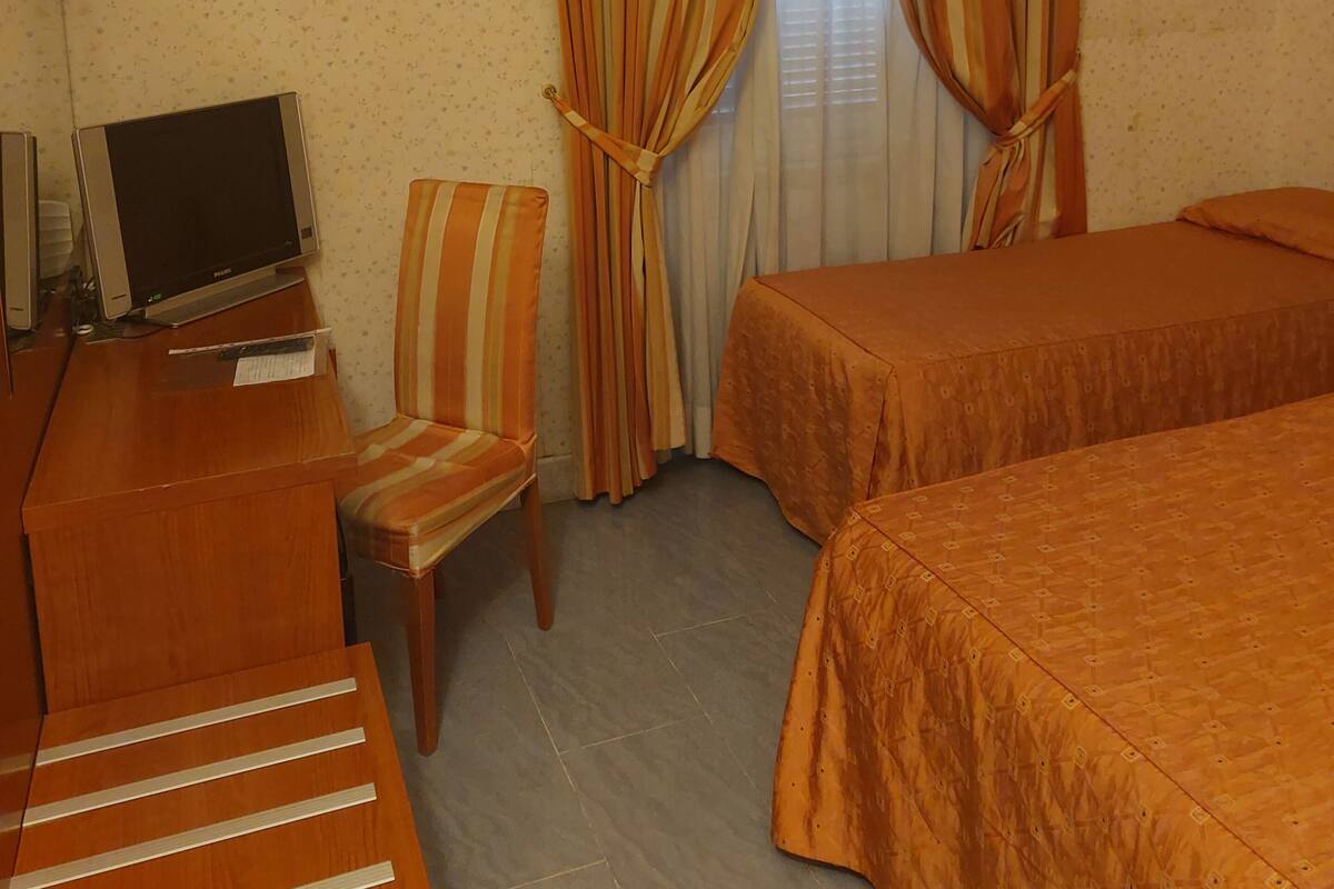 Quarto de hotel com cama, escrivaninha, computador, cadeira e cortinas.