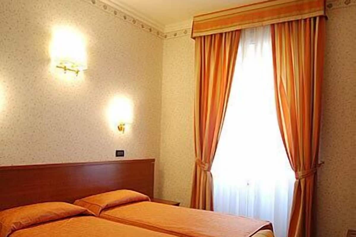Quarto de hotel com duas camas, cabeceira de madeira, luminária fixada na parede e janela com cortinas.