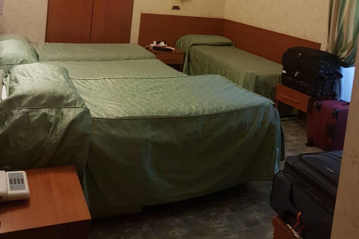 Quarto de hotel com duas camas, um guarda-roupa, uma mesa de cabeceira com abajur e uma mala.