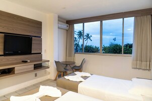 Quarto de hotel com televisão sobre uma prateleira de madeira, uma mesa pequena com duas cadeiras, cama com lençóis brancos e janela com vista para palmeiras e um edifício.