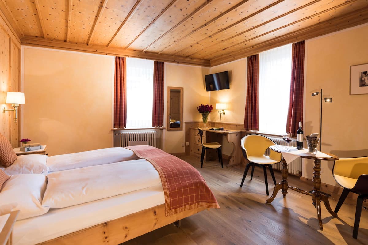 Quarto de hotel com teto de madeira, cama com manta xadrez, escrivaninha com cadeira e televisão na parede.