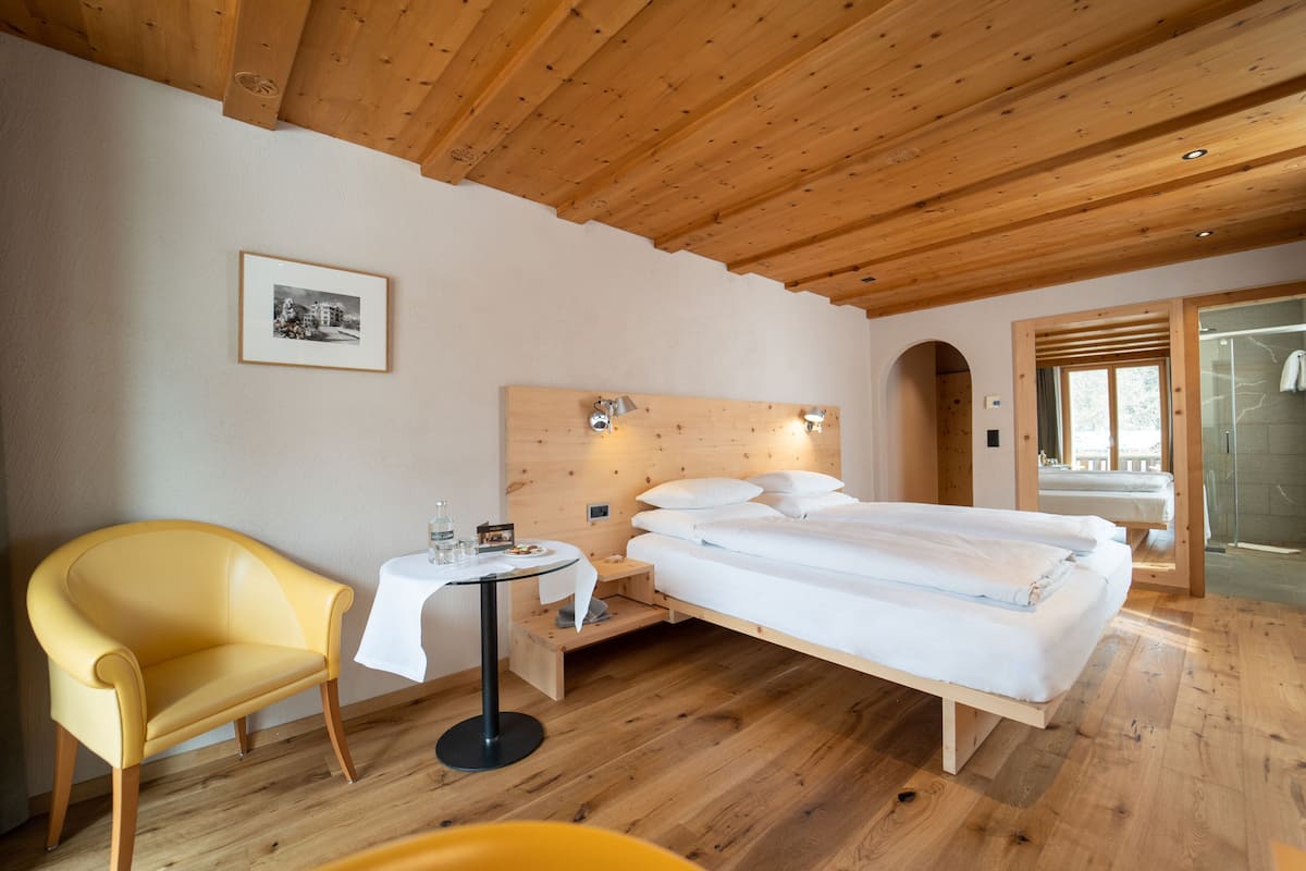Quarto moderno com teto de madeira, cama com lençóis brancos, poltrona amarela e uma pequena mesa com abajur.