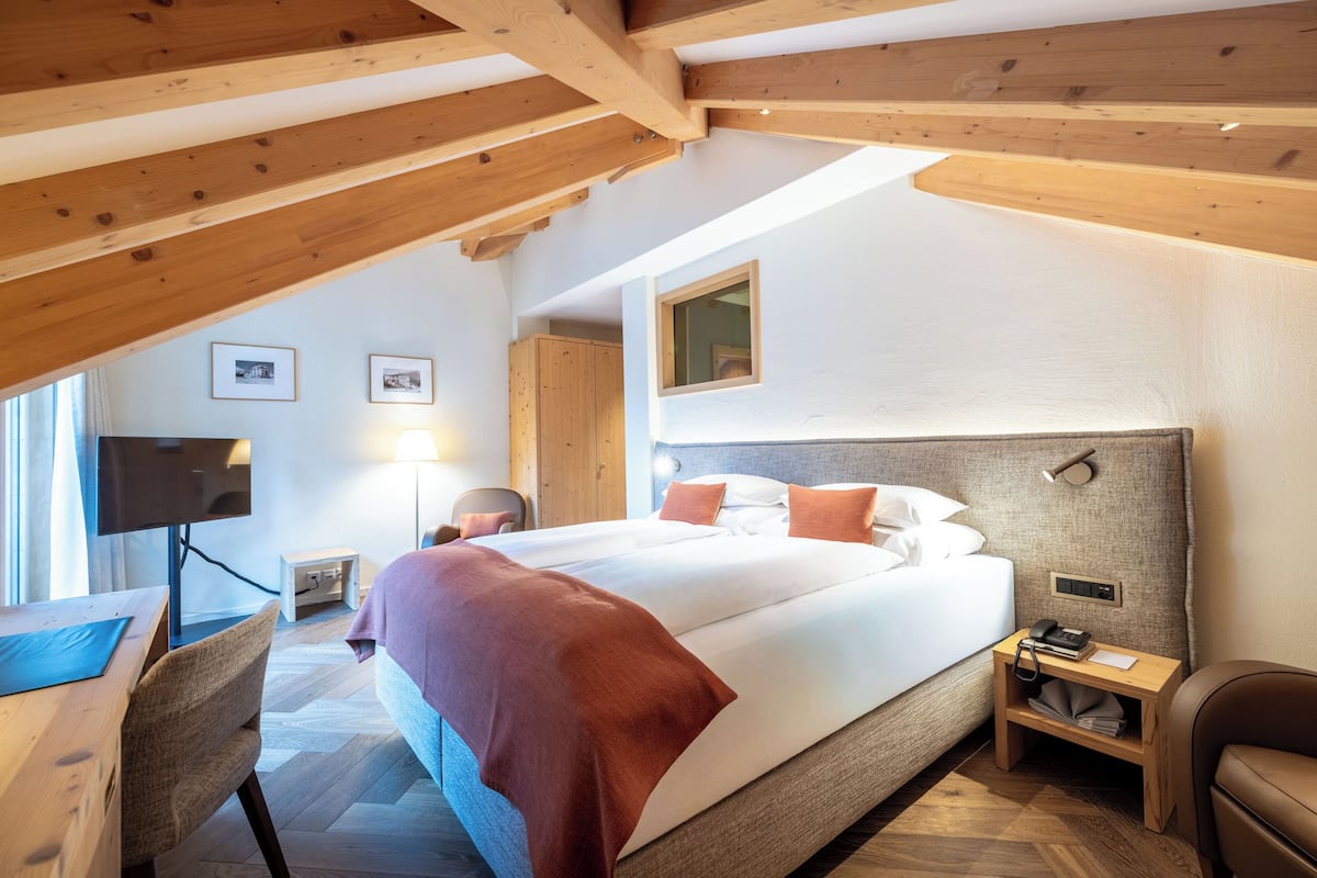Quarto com uma cama grande, vigas de madeira no teto, uma escrivaninha com cadeira, televisão e mesa de cabeceira com abajur.
