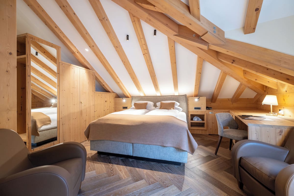 Quarto com uma cama grande, vigas de madeira no teto, uma escrivaninha e uma cadeira.