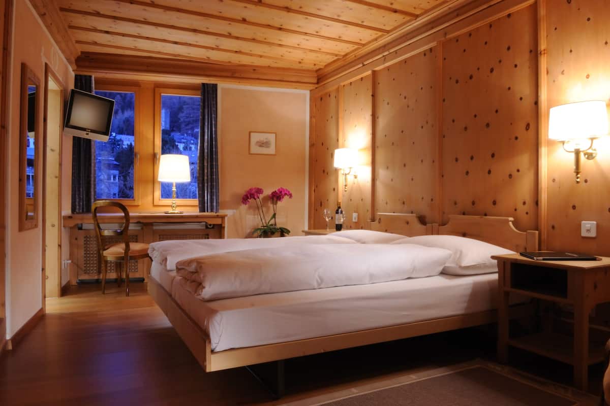 Quarto de madeira com uma cama, uma escrivaninha, uma cadeira, uma TV e duas luminárias fixadas na parede.