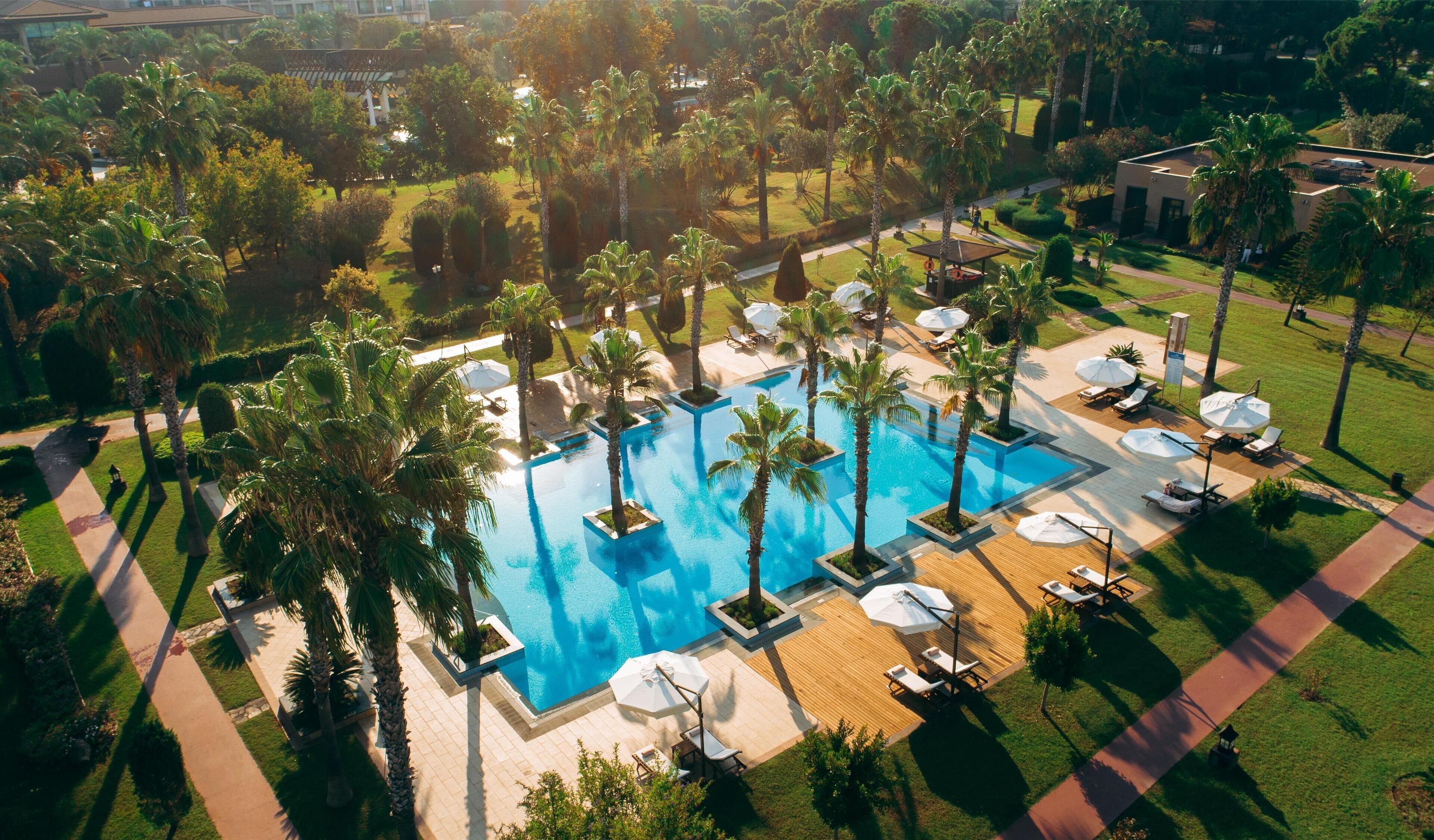 Vista aérea de um resort com uma piscina central rodeada por palmeiras, espreguiçadeiras e um deck de madeira.