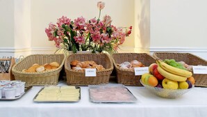 Mesa de buffet com cestas identificadas de croissants, muffins e uma saladeira de frutas.