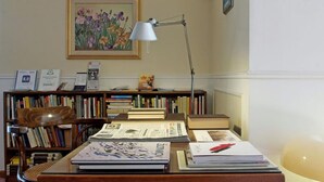 Uma sala de estudos com uma escrivaninha de madeira, estantes, uma luminária e um quadro emoldurado na parede.