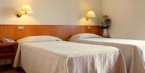 Quarto de hotel com duas camas de solteiro, cada uma com colcha branca e luminária de cabeceira. Há uma cabeceira de madeira, uma mesinha de cabeceira com telefone e um vaso com flores no lado direito.