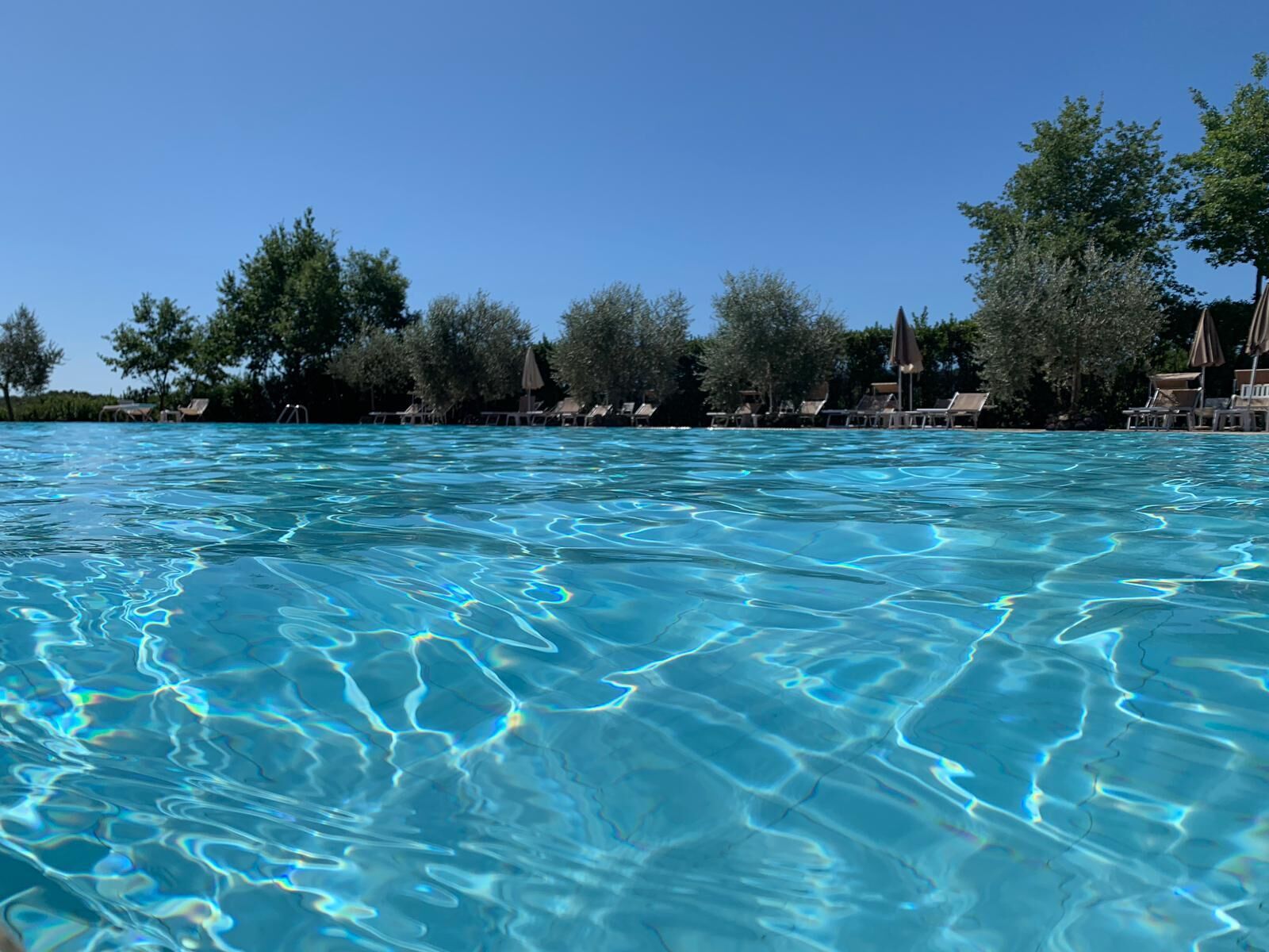 Piscina clara com ondulações visíveis, rodeada por espreguiçadeiras e guarda-sóis, indicando um ambiente de resort.