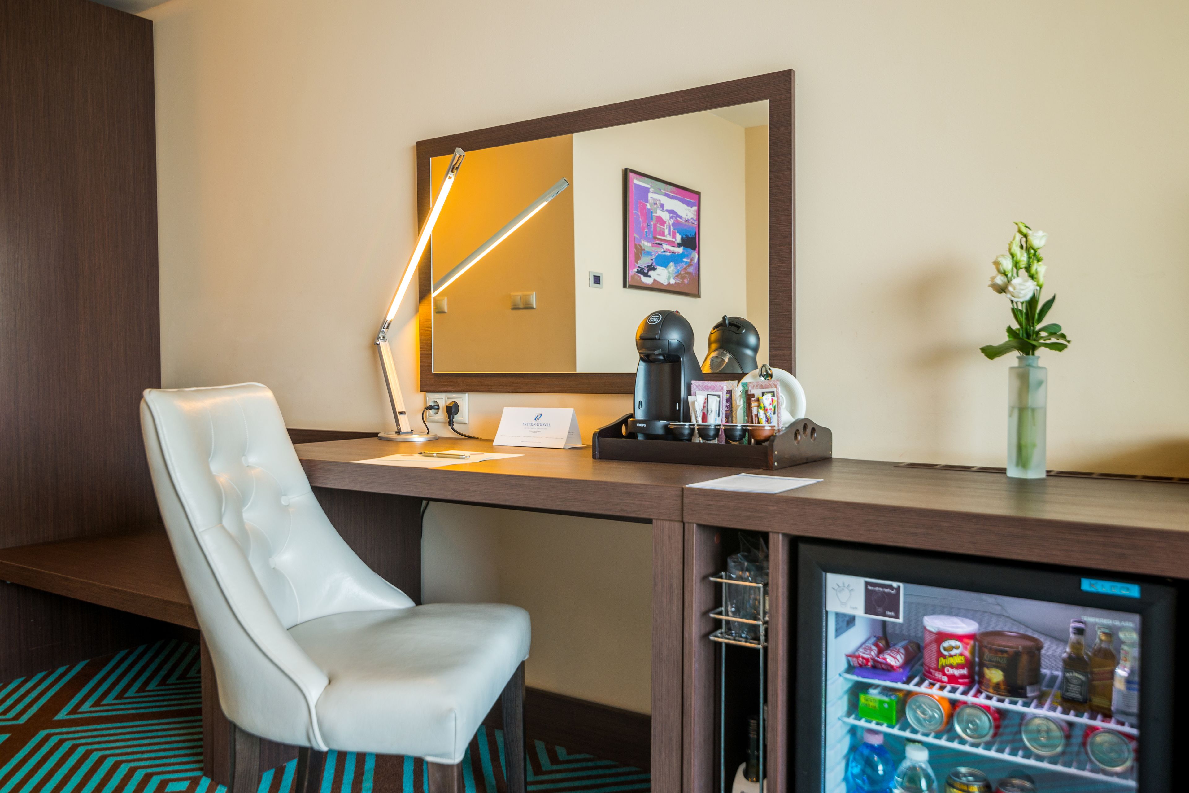 Quarto de hotel com escrivaninha, cadeira, espelho, cafeteira e mini-geladeira.