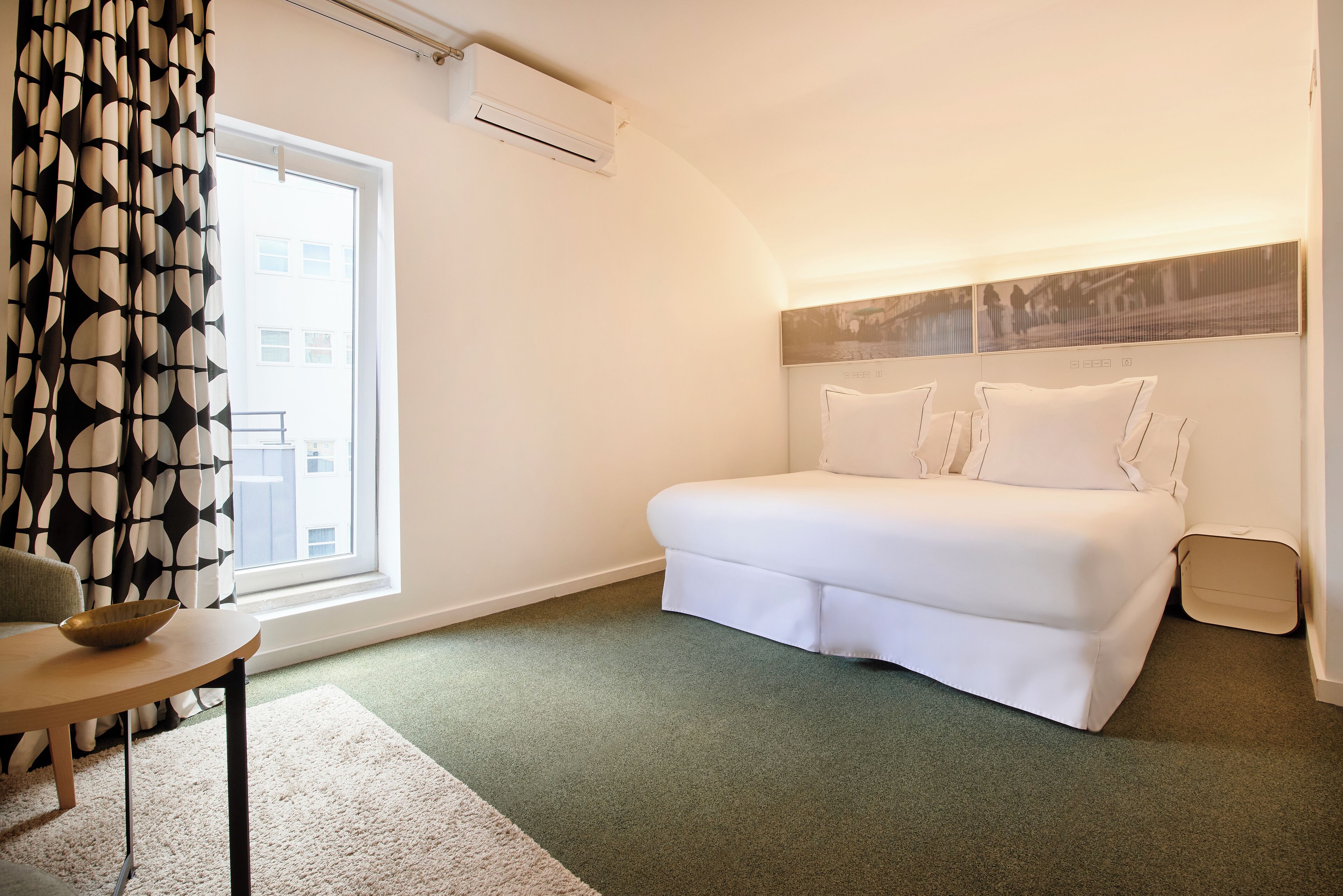 Quarto de hotel com cama, janela com cortinas, mesa pequena com um recipiente e ar condicionado fixado na parede.