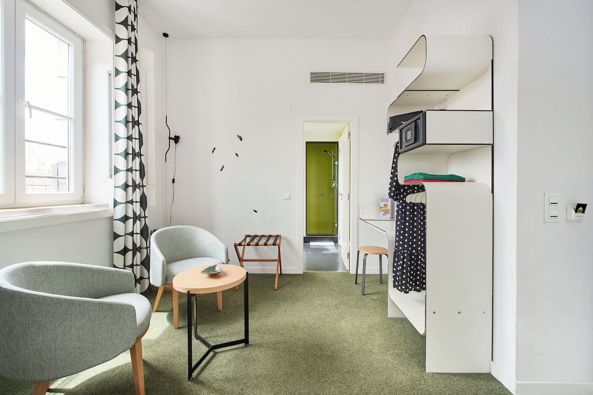 Quarto compacto com carpete verde, uma poltrona cinza, uma mesinha redonda e um guarda-roupa branco com prateleiras.