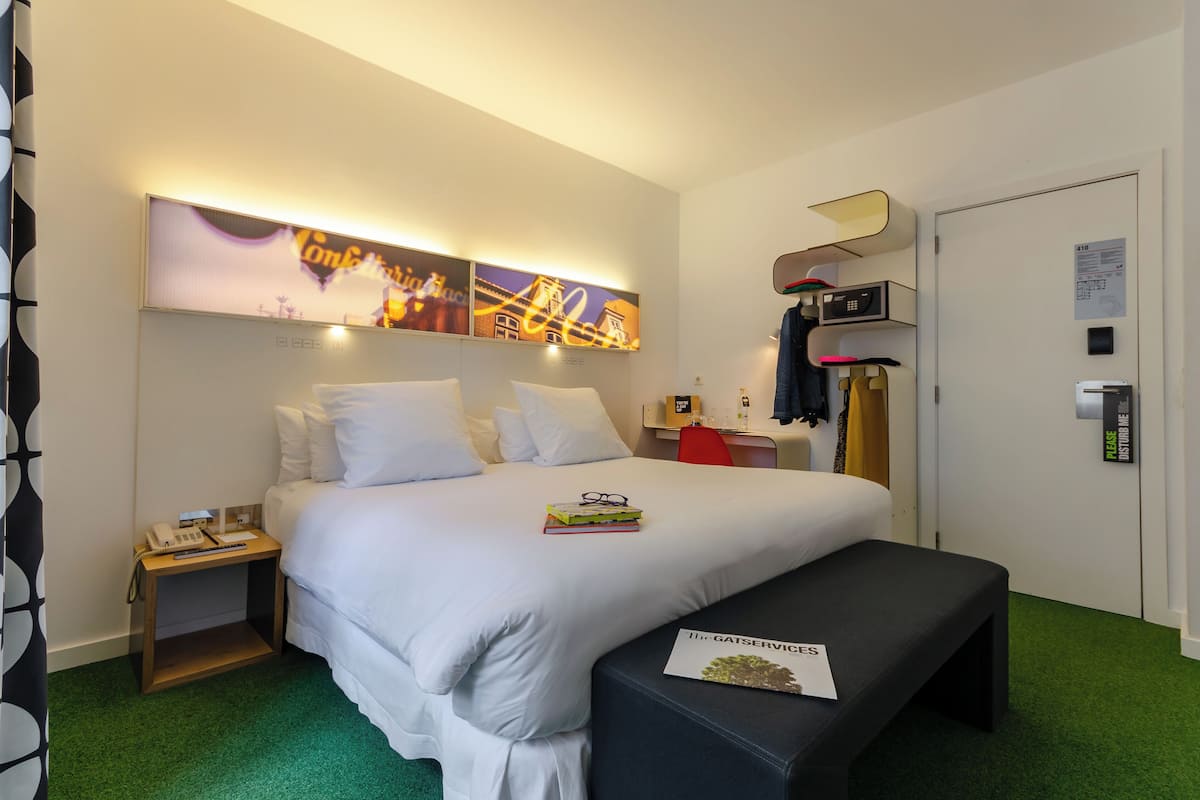 Quarto de hotel com uma cama grande, mesa de cabeceira com telefone, pequena escrivaninha com computador e um cabideiro.