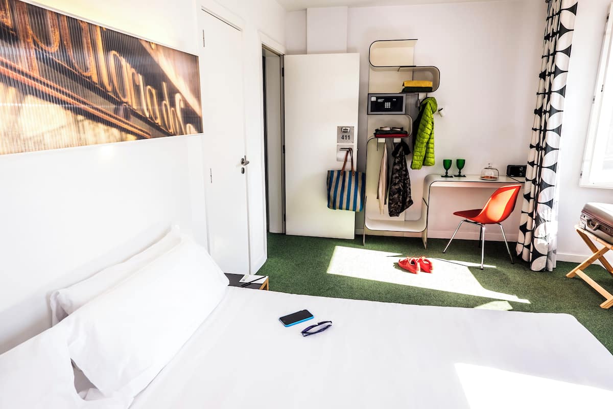 Quarto de hotel com cama, escrivaninha, cadeira, cabideiro e porta.