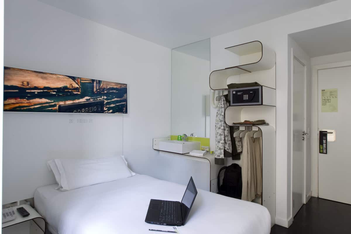 Quarto de hotel com cama, um laptop, uma pintura, um espelho, uma prateleira com artigos de higiene pessoal e uma porta.