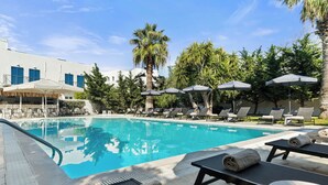 Piscina com espreguiçadeiras e guarda-sóis, cercada por palmeiras e um edifício com persianas azuis.