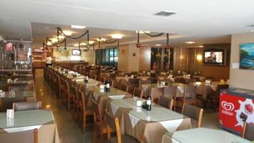 Restaurante com várias mesas, cadeiras, um bar e uma tela de televisão.