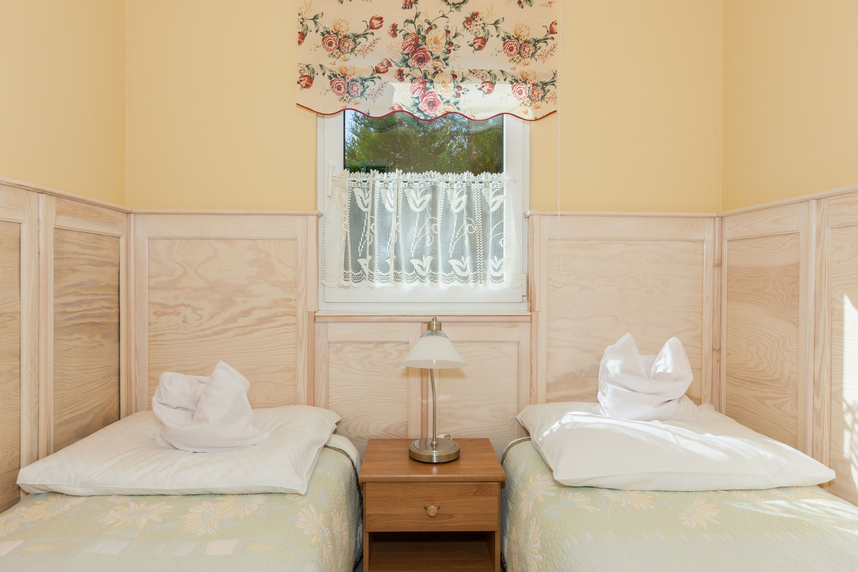Quarto com duas camas, mesa de cabeceira com abajur, cortinas florais e janela com vista para árvores.