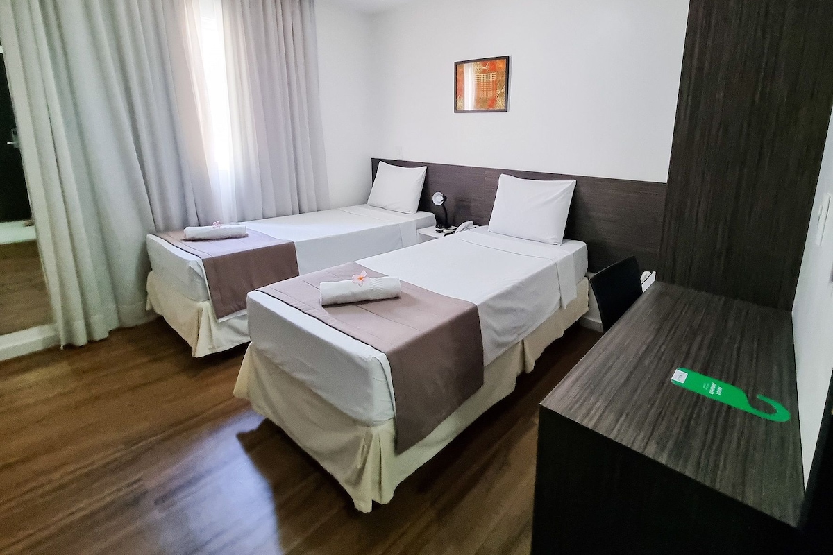 Quarto de hotel com duas camas de solteiro, uma escrivaninha, uma cadeira, um espelho e uma janela com cortinas.