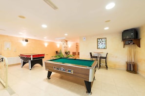Uma mesa de bilhar, uma mesa de futebol de mesa e uma televisão num espaço com paredes bege e chão em azulejos.