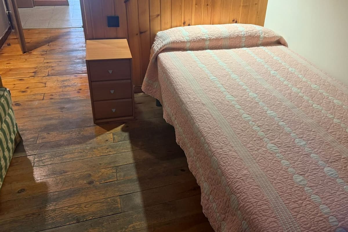 Quarto com piso de madeira, uma cama de solteiro com colcha estampada, mesa de cabeceira e cômoda.