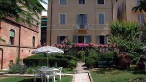 Um edifício de dois andares com entrada central, um pequeno jardim com mesa e cadeiras, e um caminho que leva à entrada.