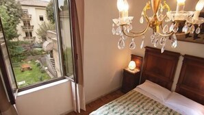 Quarto com uma grande janela, um candelabro, uma mesa de cabeceira com abajur e uma cama com colcha estampada.