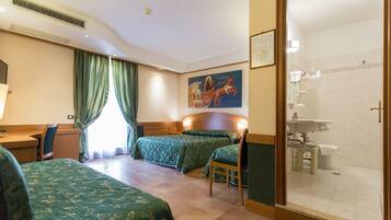 Quarto de hotel com duas camas, uma escrivaninha, uma cadeira, um quadro na parede e um banheiro visível por uma porta aberta.