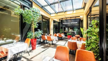 Um átrio de restaurante com mesas preparadas para refeições, grandes vasos de plantas e teto de vidro.