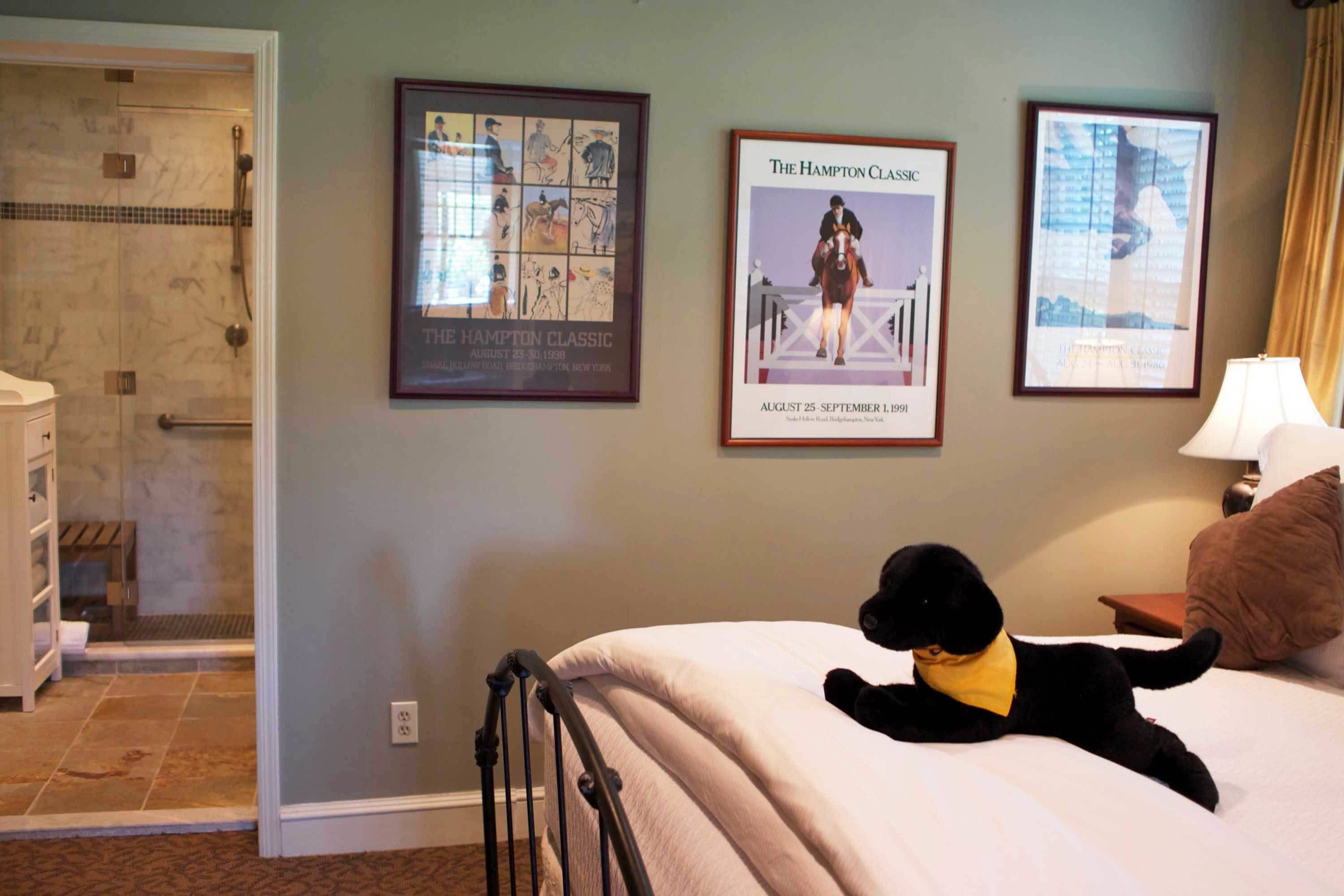Quarto de hotel com uma cama, um quadro com o cartaz de 'The Hampton Classic', área de chuveiro e uma pequena mesa com abajur.