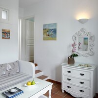 Quarto com sofá branco, mesa de vidro com frutas, cômoda branca com gavetas e espelho fixado na parede.