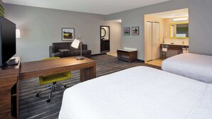 Quarto de hotel com cama, escrivaninha, cadeira, televisão, luminária e banheiro com espelho e pia.