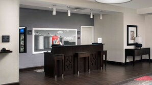 Uma área de recepção de hotel com design moderno, apresentando um balcão de recepção, uma tela digital exibindo o nome do hotel e uma grande obra de arte emoldurada na parede.