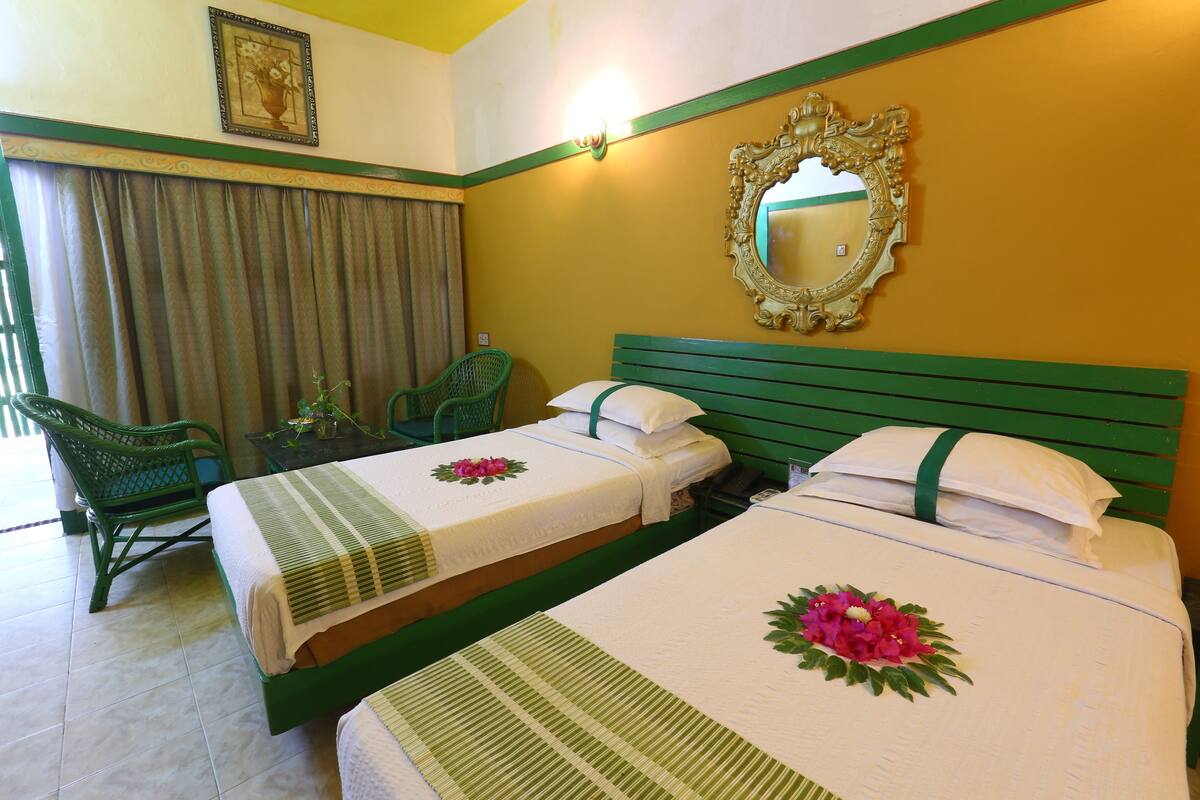 Quarto de hotel com duas camas, cabeceiras verdes, um espelho, uma cadeira verde e uma pequena mesa com uma planta.