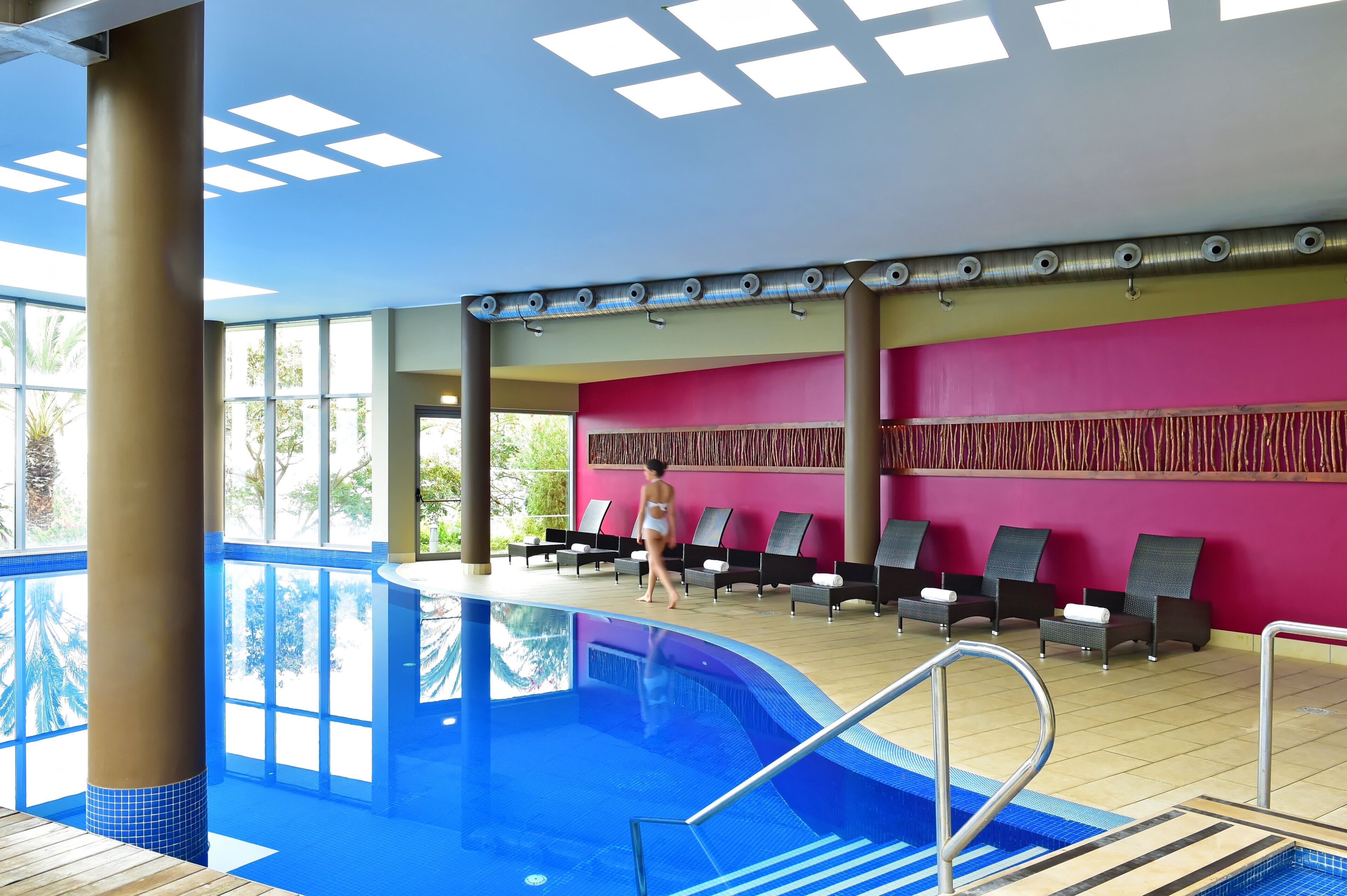 Piscina interior com vista clara da área da piscina, espreguiçadeiras e uma pessoa caminhando.