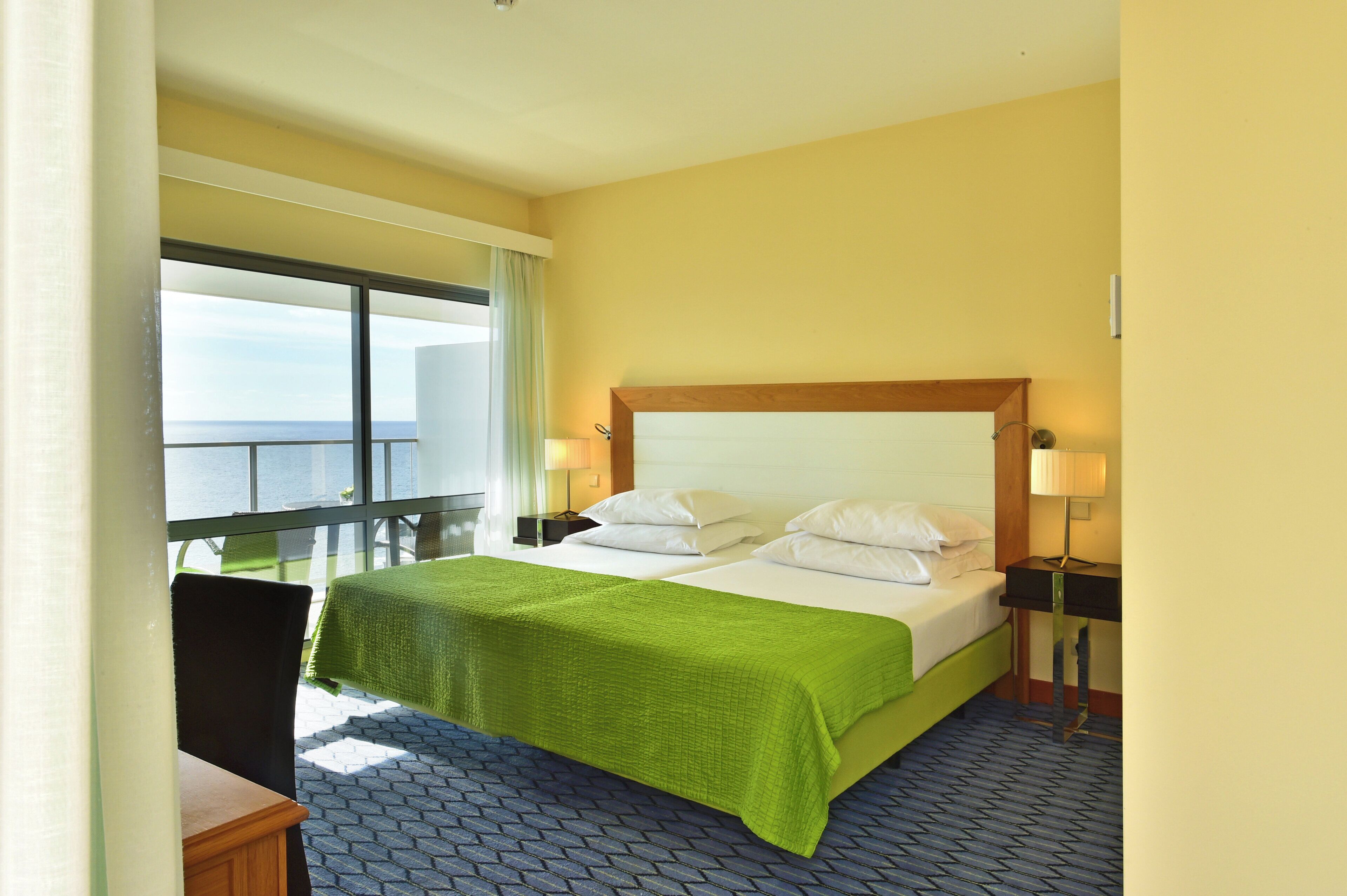 Quarto de hotel com uma cama, uma cadeira, uma escrivaninha e vista para o mar através de uma grande janela.
