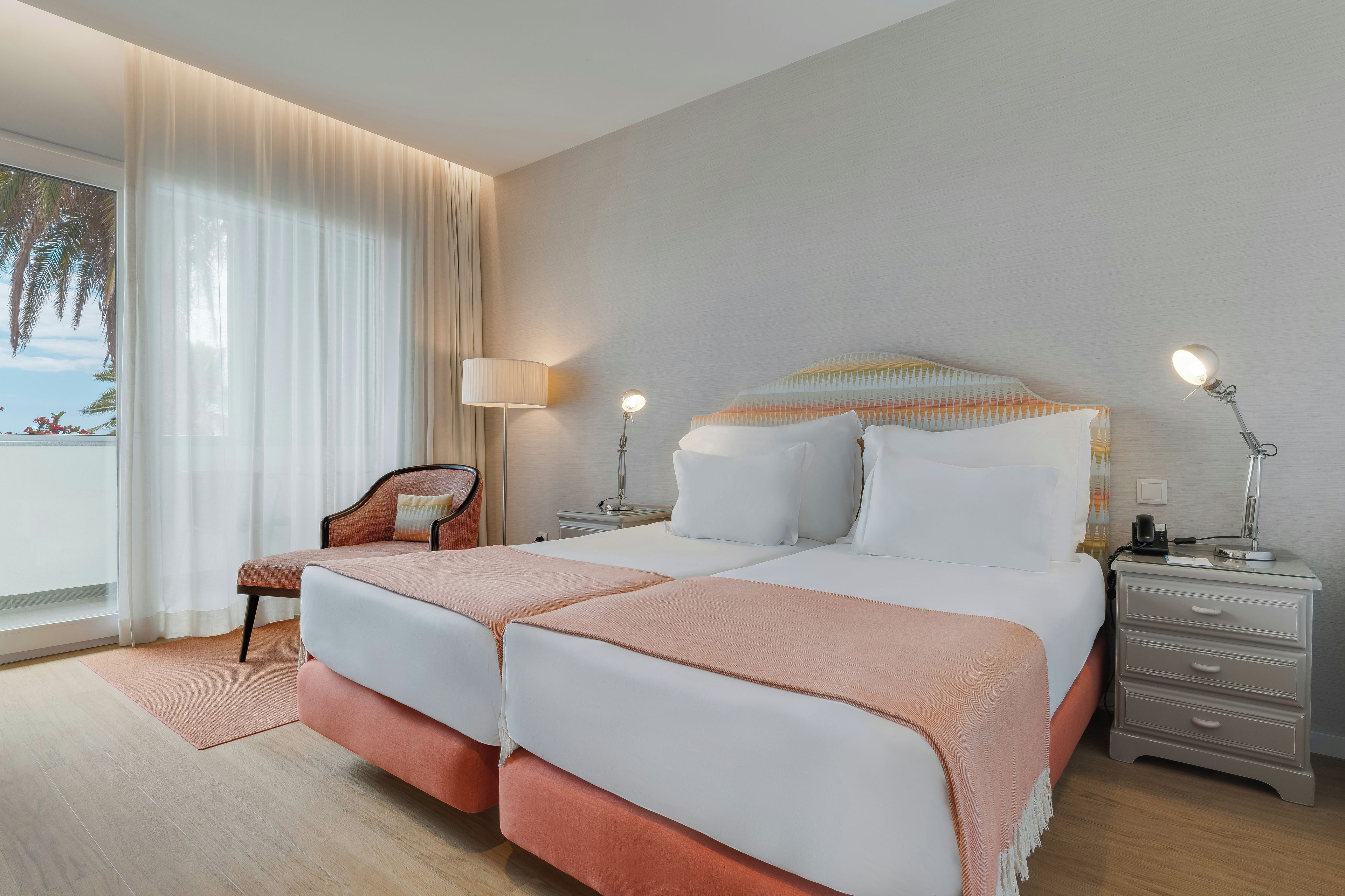 Quarto de hotel com uma cama, duas mesinhas de cabeceira, uma cadeira, uma luminária e vista para palmeiras.