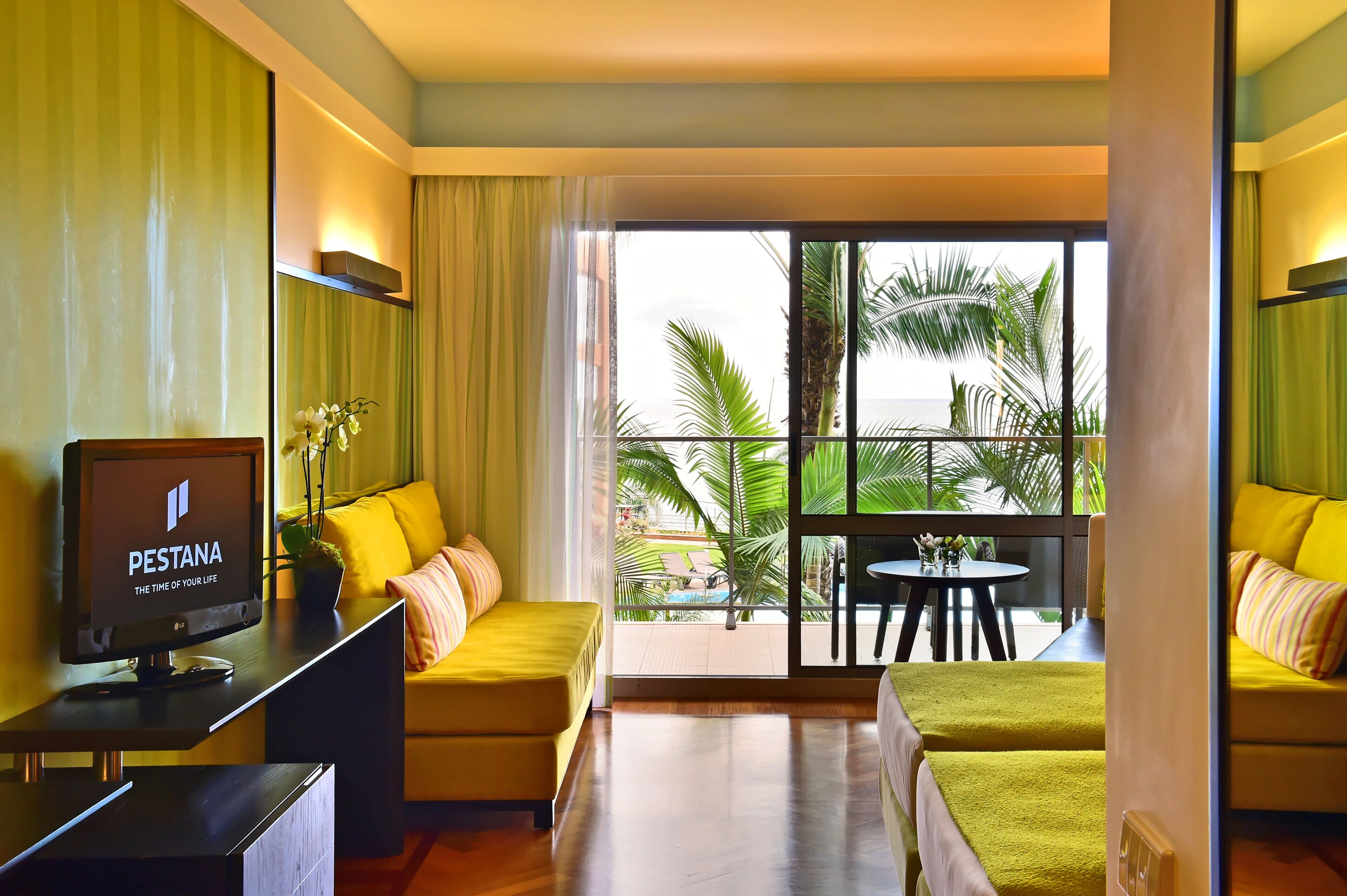 Quarto de hotel com uma grande janela, um sofá, uma TV e uma pequena mesa com um vaso de flores.